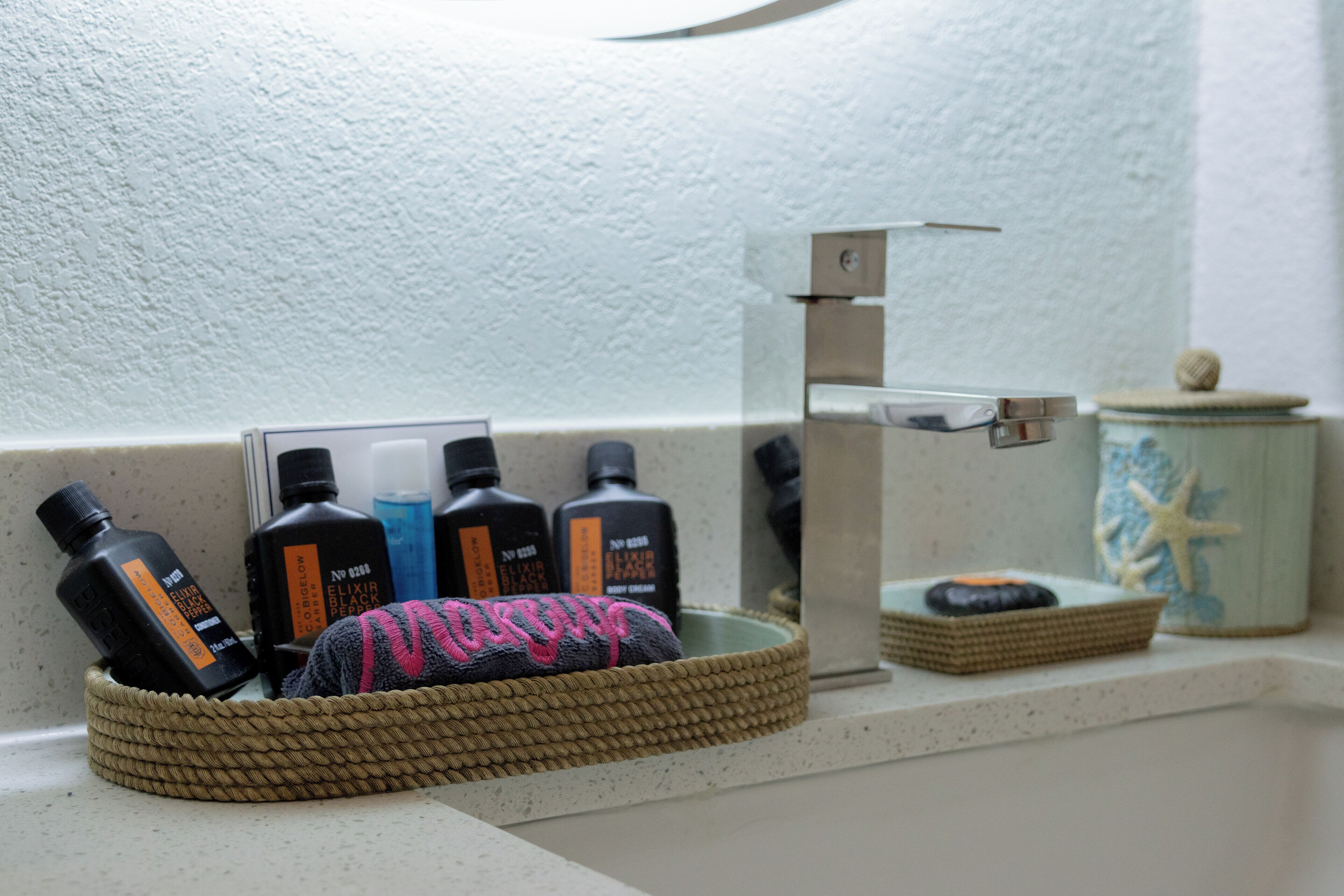 Casa de banho com cesto de fibra natural, dispensador de sabonete em vidro e recipiente cerâmico decorativo.