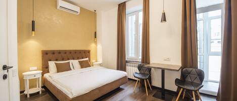 Quarto de hotel moderno com uma cama grande, uma mesa de trabalho e uma cadeira. Há uma janela com cortinas, um aparelho de ar condicionado e um lustre.