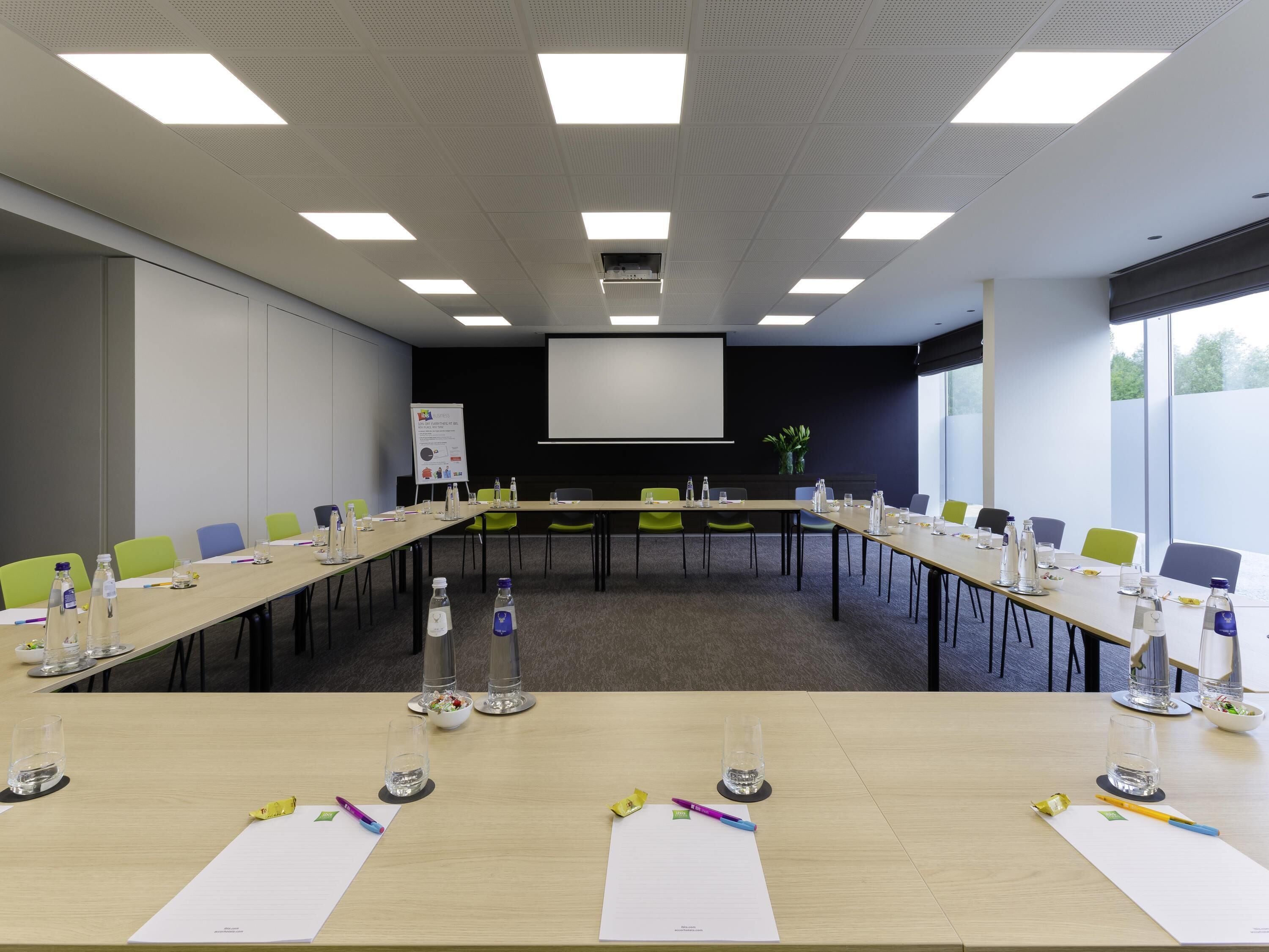 Uma sala de conferências com uma mesa comprida, cadeiras, uma tela de projetor e um quadro branco.