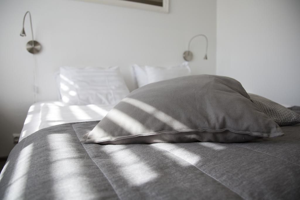Uma cama com cobertor cinza e travesseiros, parede branca e luminária fixada na parede.