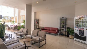 Um hall amplo com um sofá vermelho, várias poltronas, uma máquina de café e plantas em vasos.