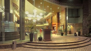 Um hall de hotel moderno com fachada de vidro, escadaria central e plantas em vasos.