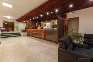 Uma área de recepção de hotel com um balcão de madeira, um recepcionista e um espaço de estar com um sofá e uma planta em vaso.