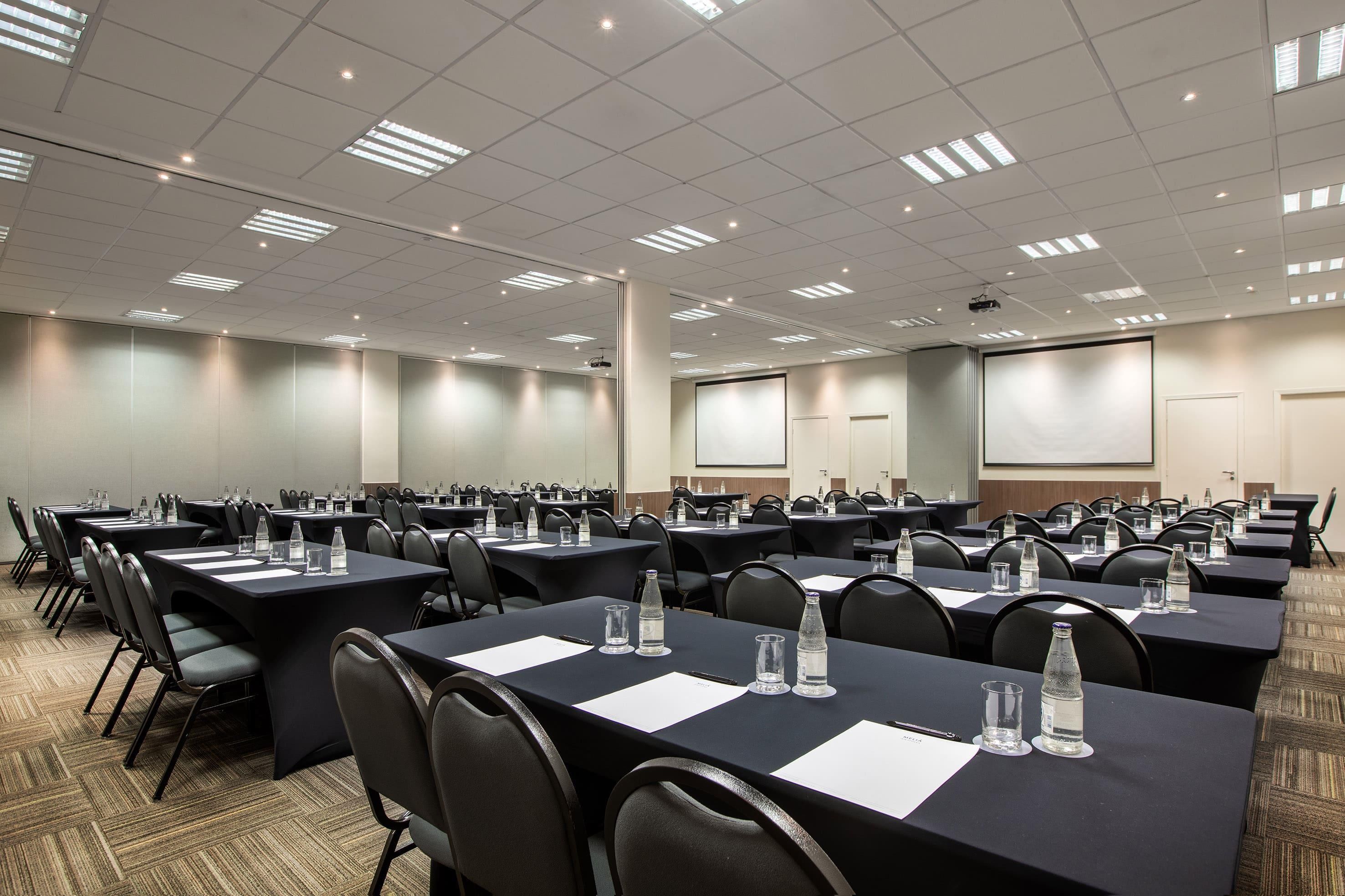 Sala de conferências com várias mesas, cadeiras e telas de apresentação.