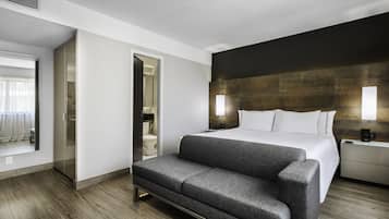 Quarto de hotel moderno com uma cama grande, um sofá, um closet espaçoso e um banheiro visível através de uma porta aberta.