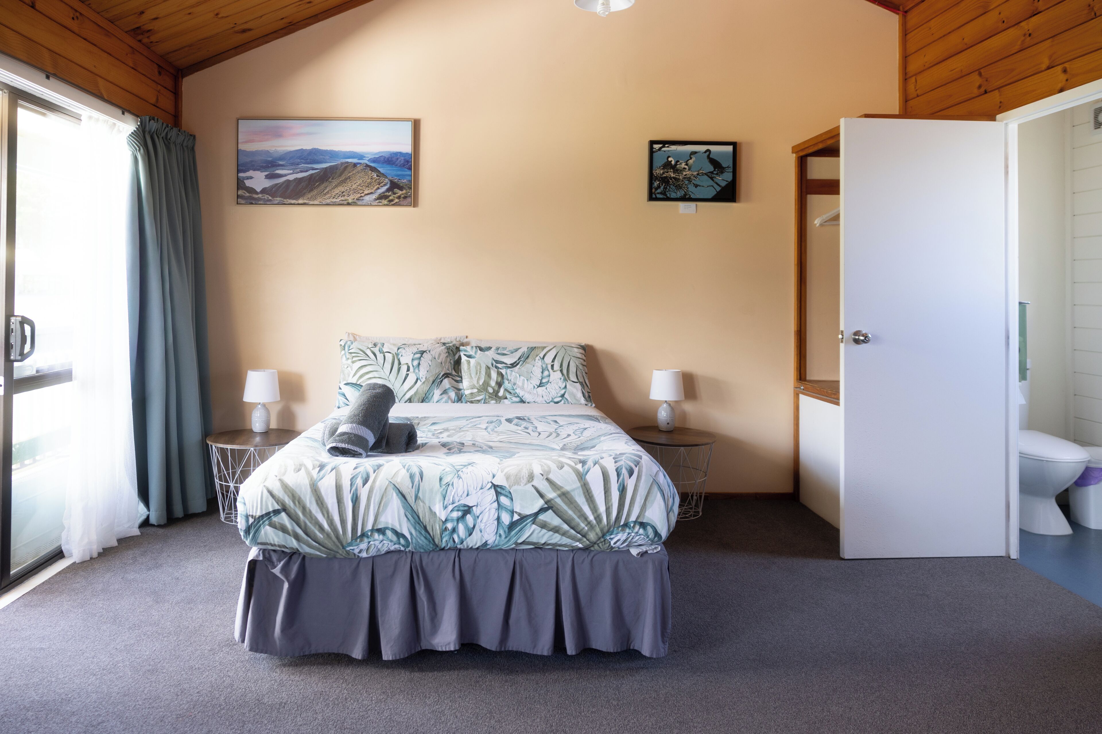 Um quarto com uma cama, duas mesas de cabeceira, uma janela com cortinas, uma porta e uma parede com duas imagens emolduradas.