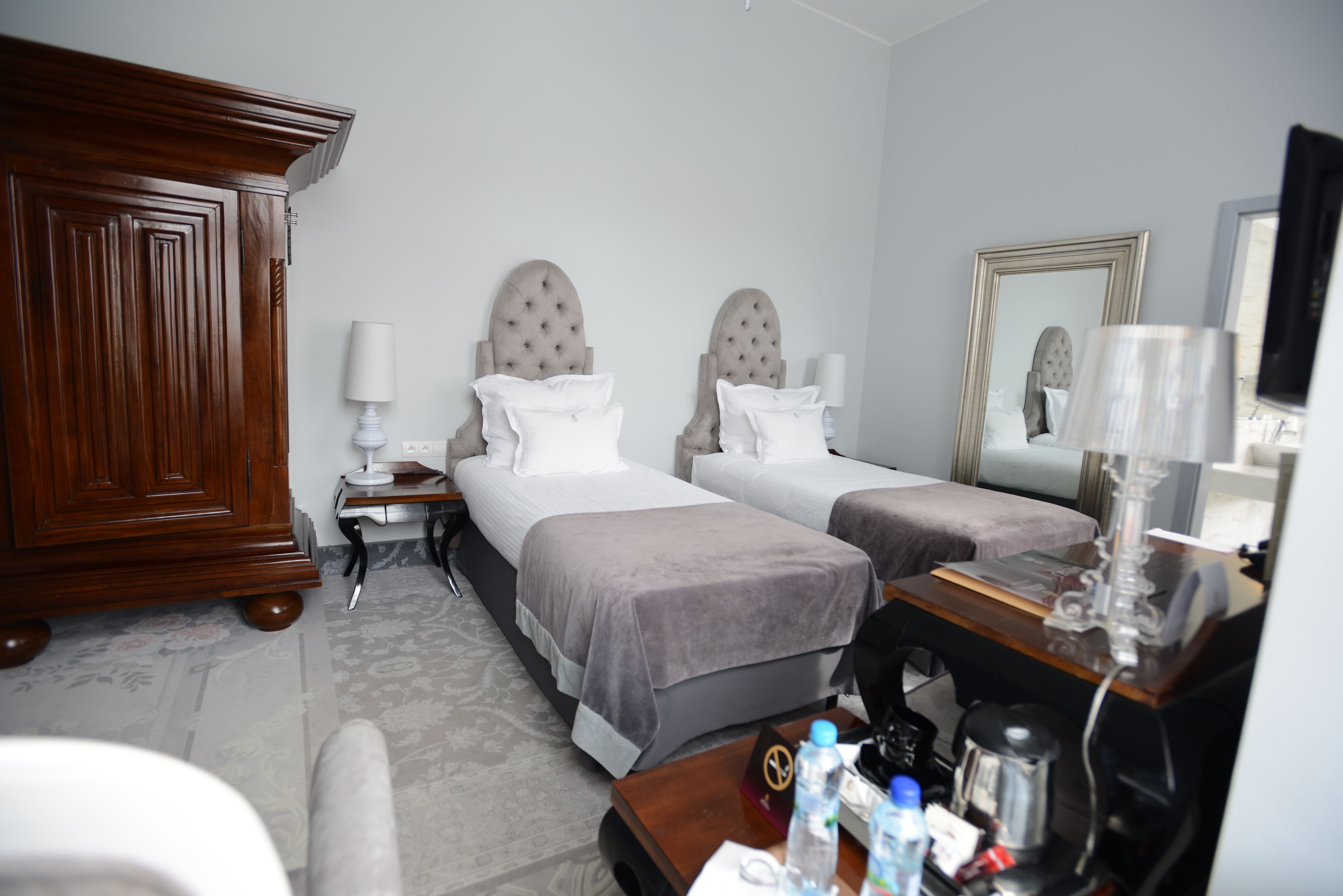 Quarto de hotel com duas camas, guarda-roupa de madeira, secretária com abajur e espelho.