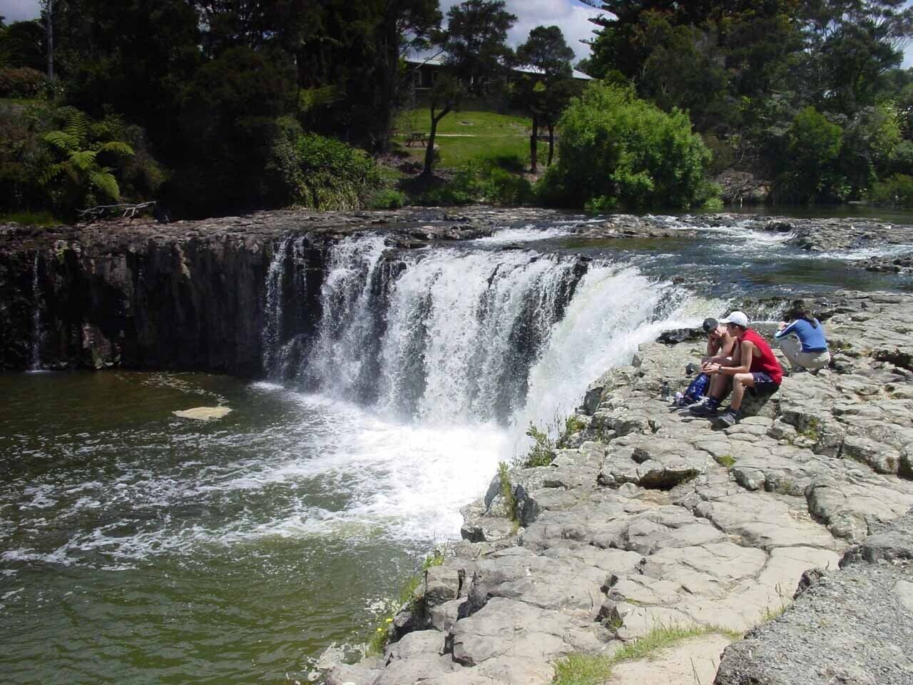 Uma cascata deságua sobre um afloramento rochoso em um rio, com pessoas sentadas nas pedras por perto.