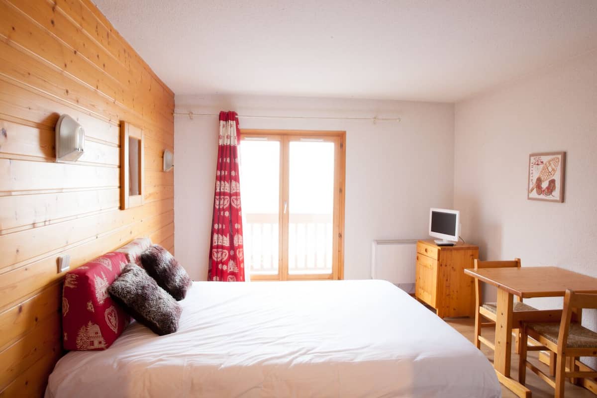 Quarto com cabeceira de madeira, cama com roupa de cama branca, cortina vermelha estampada, televisão sobre um armário de madeira e uma pequena mesa com cadeiras.