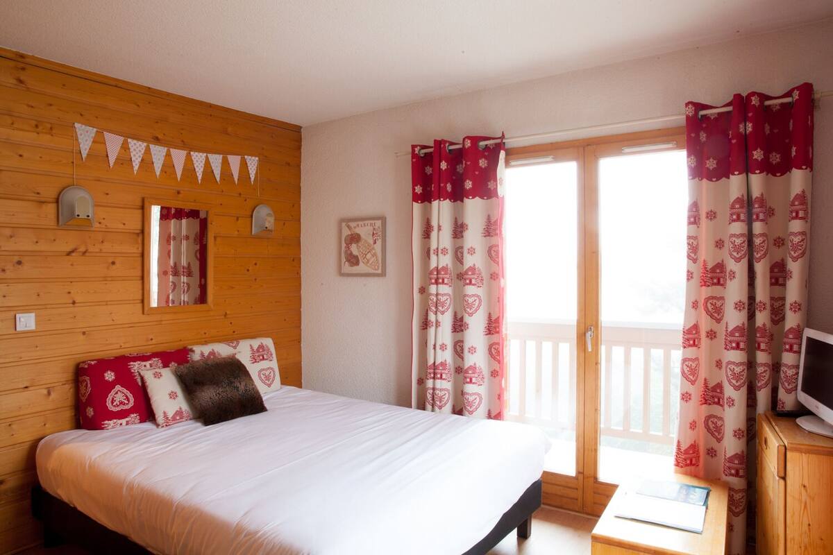 Quarto com cabeceira de madeira, cama com roupa de cama branca, cortinas estampadas em vermelho e branco, e uma televisão sobre um armário de madeira.