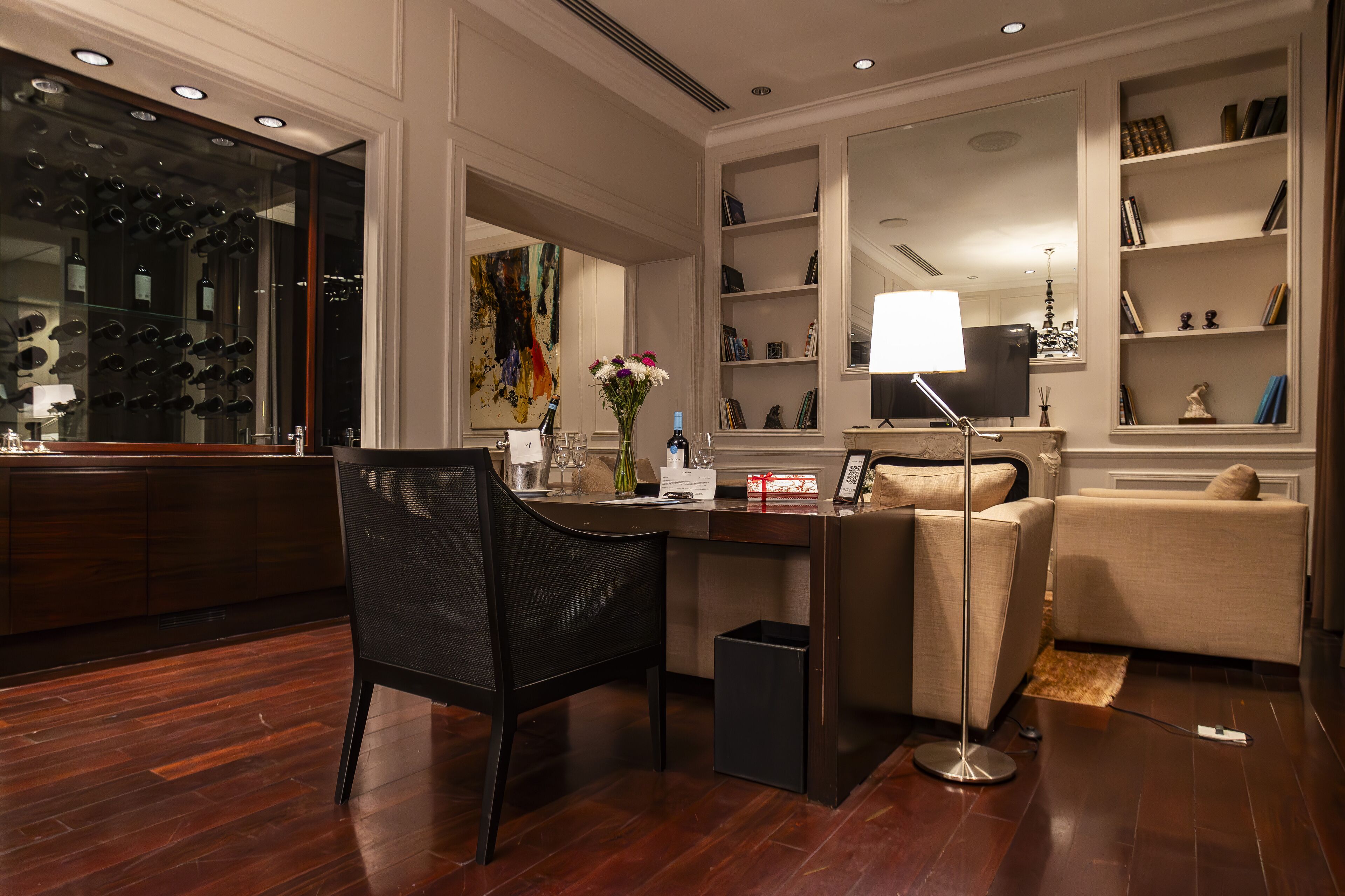 Quarto de hotel moderno com piso de madeira, uma escrivaninha com cadeira, um sofá, uma estante e um espelho grande.