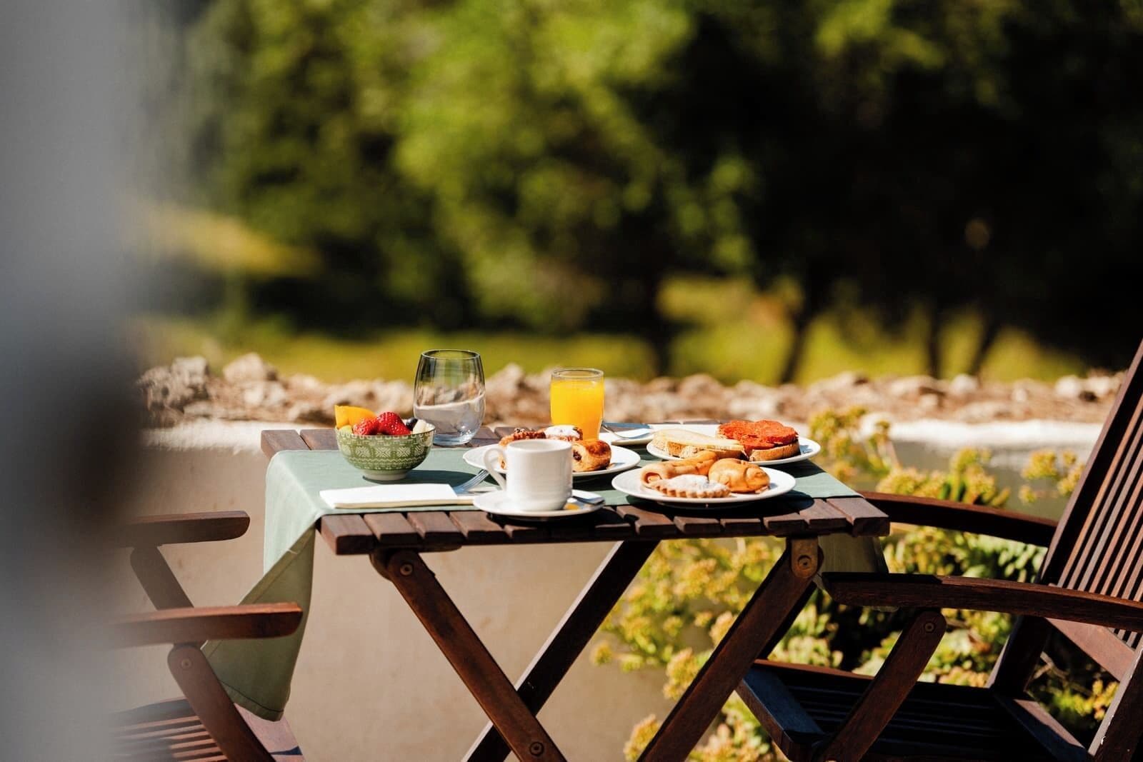 Mesa de jantar montada ao ar livre com itens de café da manhã, incluindo frutas, pão e um copo de suco de laranja.