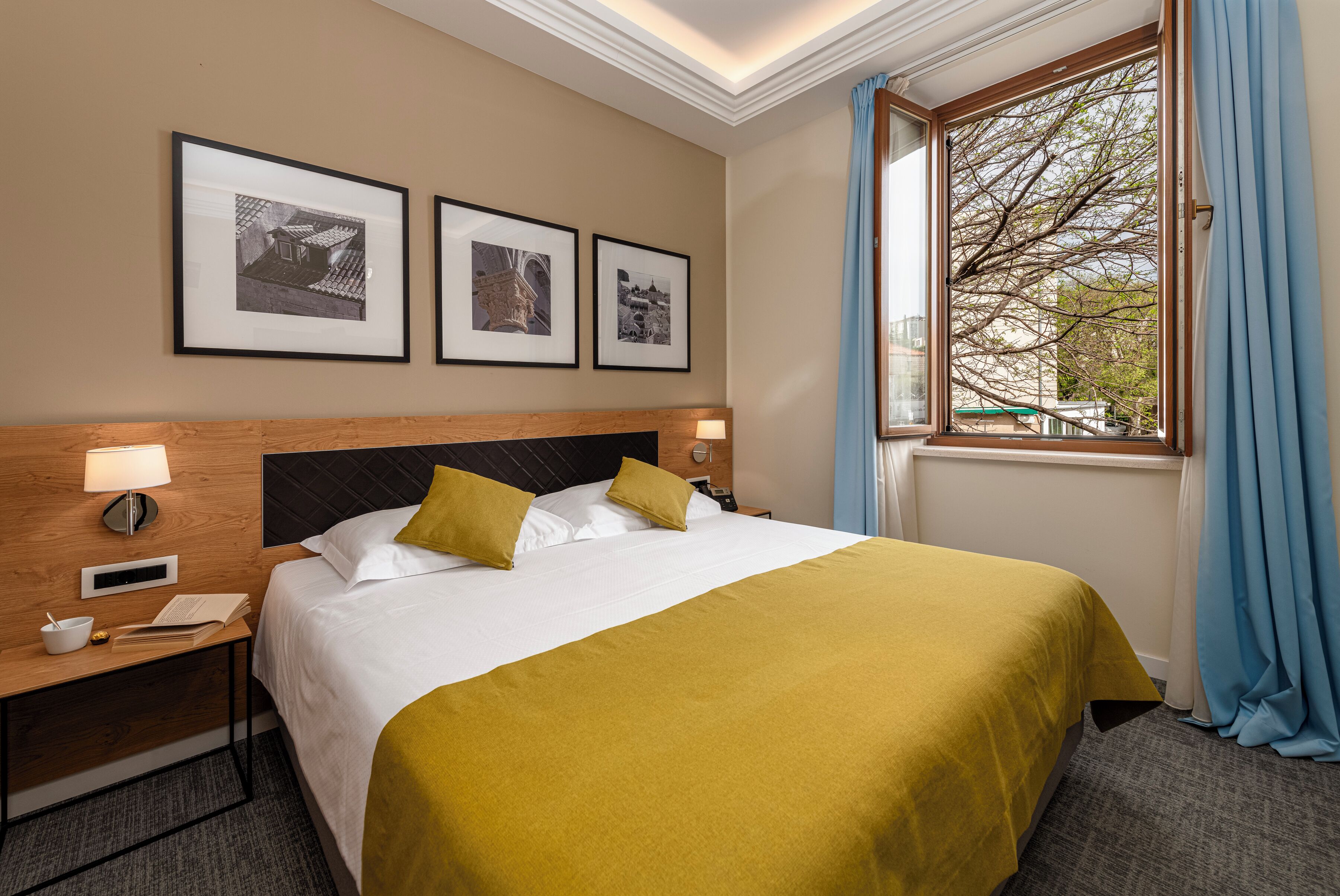 Quarto de hotel com cama, mesa de cabeceira, luminária, janela com cortinas e três quadros na parede.