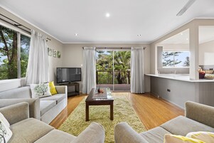 Uma sala de estar moderna com um sofá, poltronas, uma mesa de centro e uma TV. Há uma porta de vidro deslizante que dá para uma varanda com vista para o verde.
