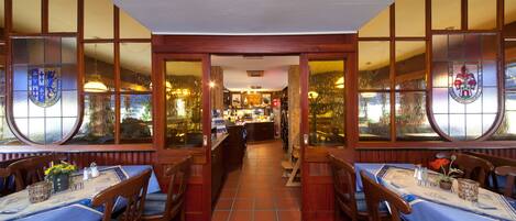 Um restaurante com mobiliário de madeira, chão ladrilhado e grandes janelas com painéis de vidro decorativos.