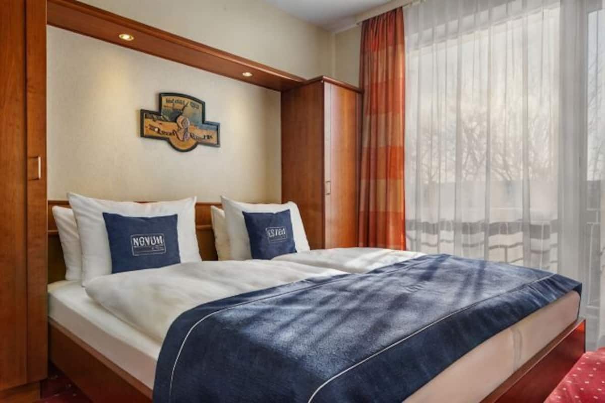 Quarto de hotel com cama, cabeceira, decoração na parede e janela com cortinas.
