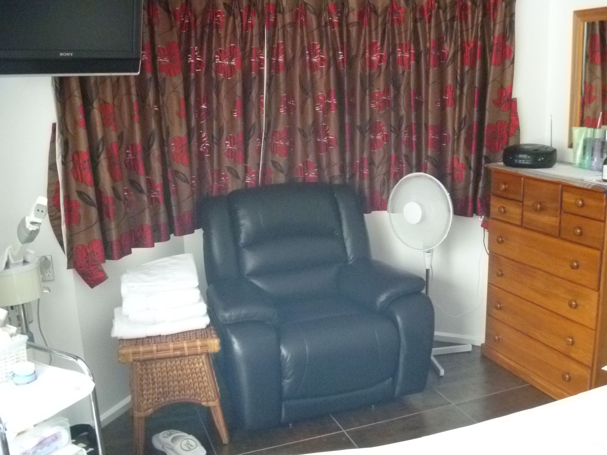 Quarto com cadeira de couro preto, ventilador de chão, cômoda de madeira e televisão na parede.
