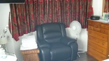 Quarto com cadeira de couro preto, ventilador de chão, cômoda de madeira e televisão na parede.