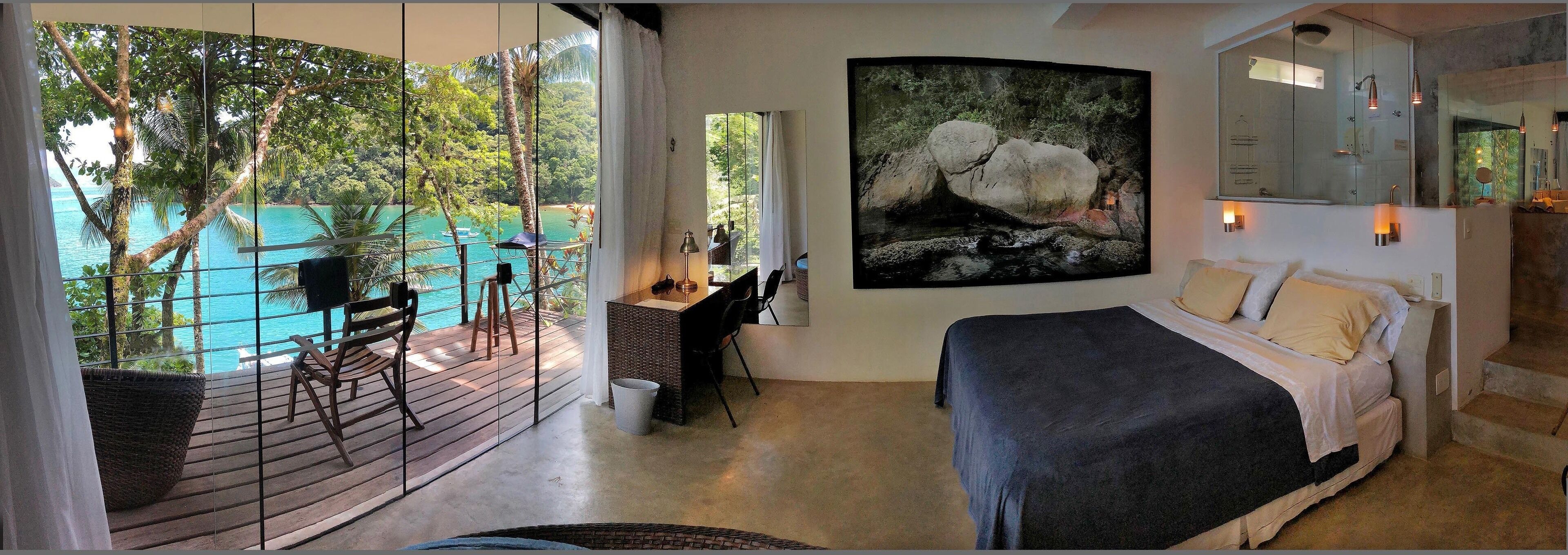 Quarto com cama, escrivaninha e cadeira, uma janela ampla com vista para uma piscina e árvores, e um quadro de pedras na parede.