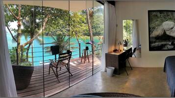 Quarto com cama, escrivaninha e cadeira, uma janela ampla com vista para uma piscina e árvores, e um quadro de pedras na parede.