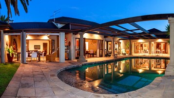 Uma área junto à piscina com um alpendre coberto, espaço para refeições e vista para a casa.