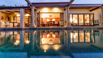 Vista da piscina de um resort, com área de estar coberta, espaço para refeições e reflexo na água.
