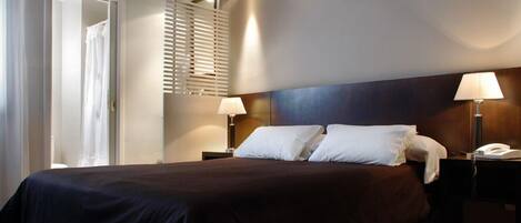 Quarto de hotel com uma cama grande, duas luminárias de cabeceira, um painel de madeira na cabeceira e uma porta que dá para um banheiro.