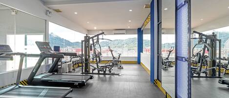 Academia com esteiras, aparelhos de musculação e uma grande janela com vista para uma paisagem montanhosa.