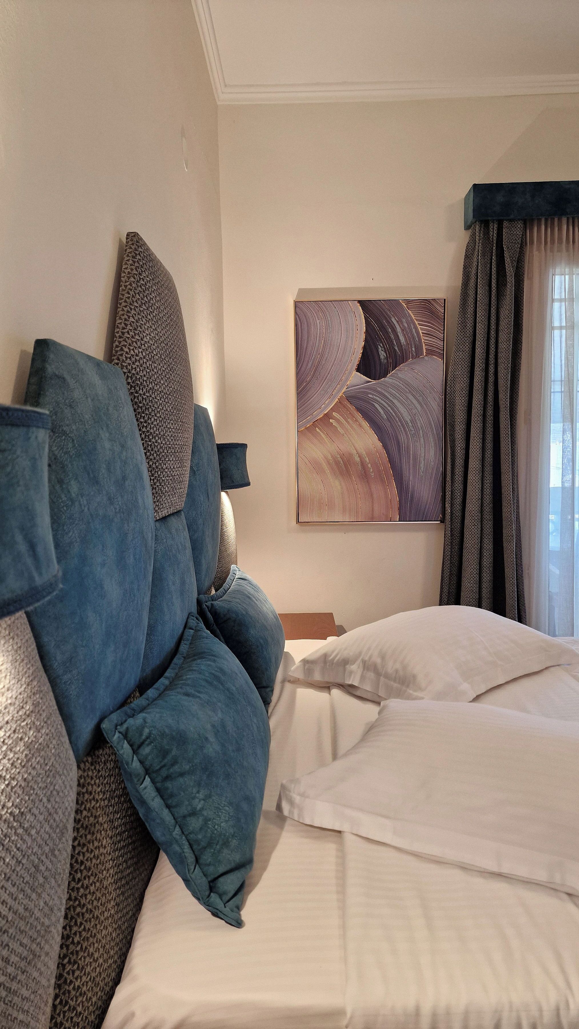Quarto de hotel com uma cama grande, cabeceiras azuis, um quadro na parede e uma janela com cortinas.