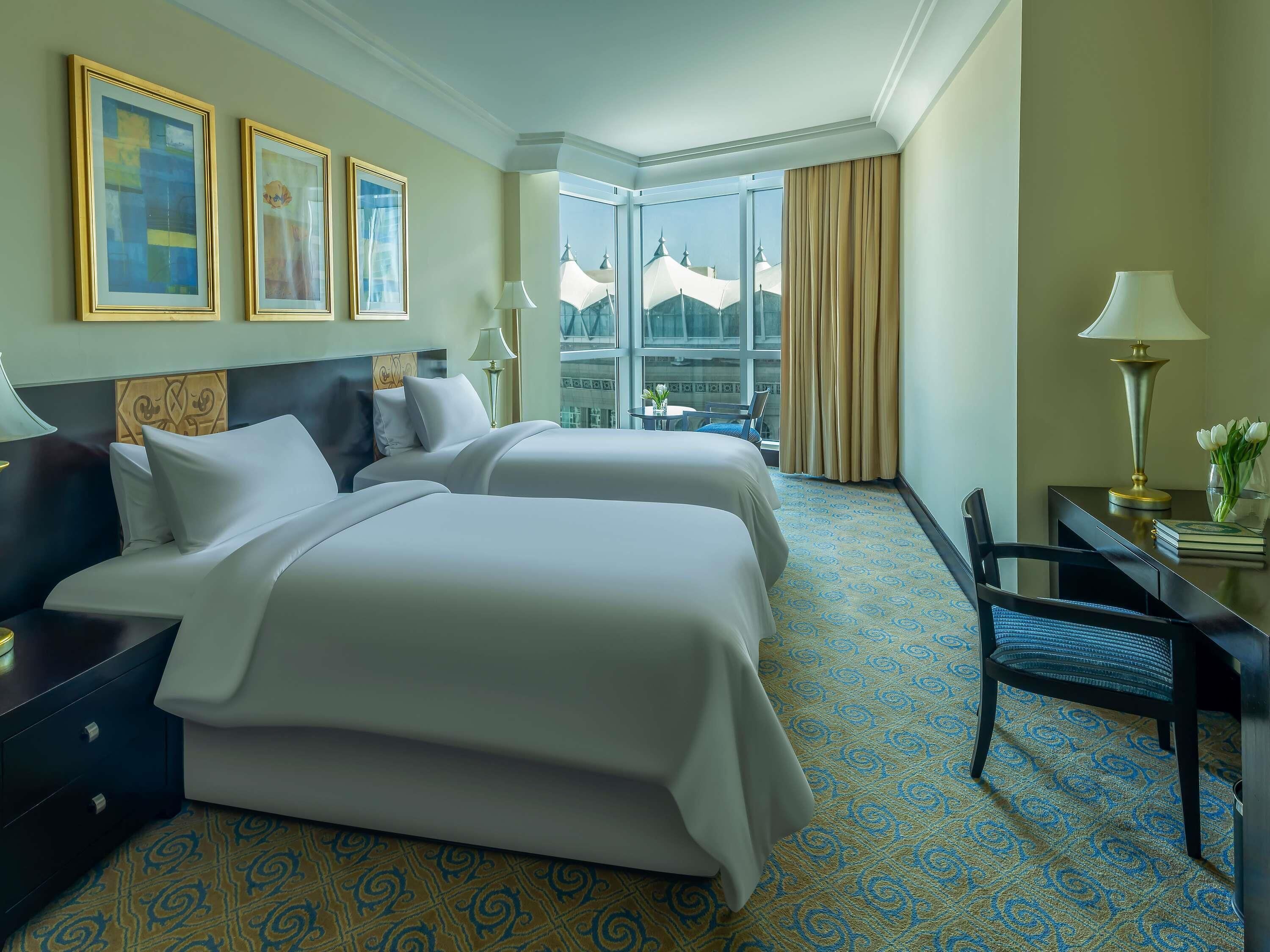 Quarto de hotel com duas camas, uma mesa com luminária, uma cadeira e vista para um edifício com tendas brancas.
