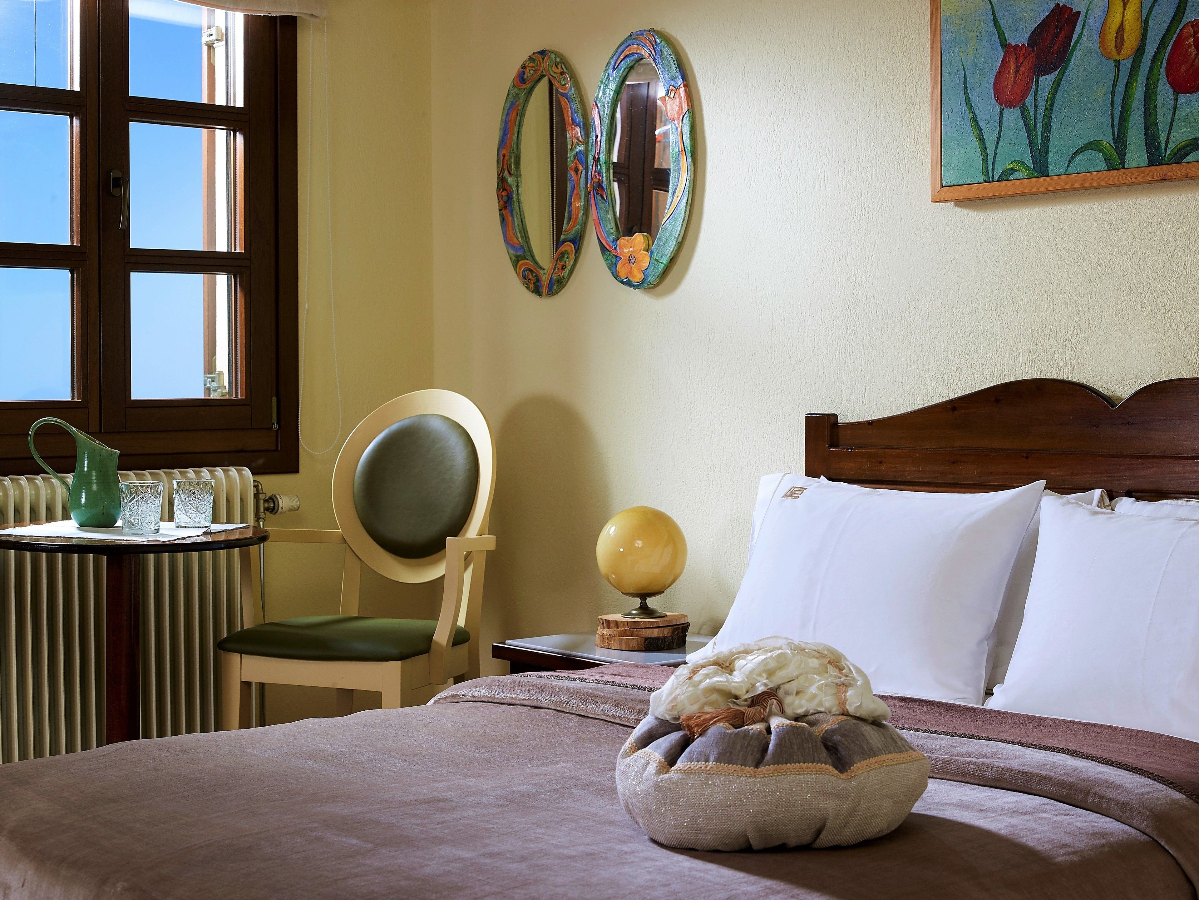 Quarto com cama, uma cadeira, mesa com jarra e copos, radiador e dois espelhos na parede.