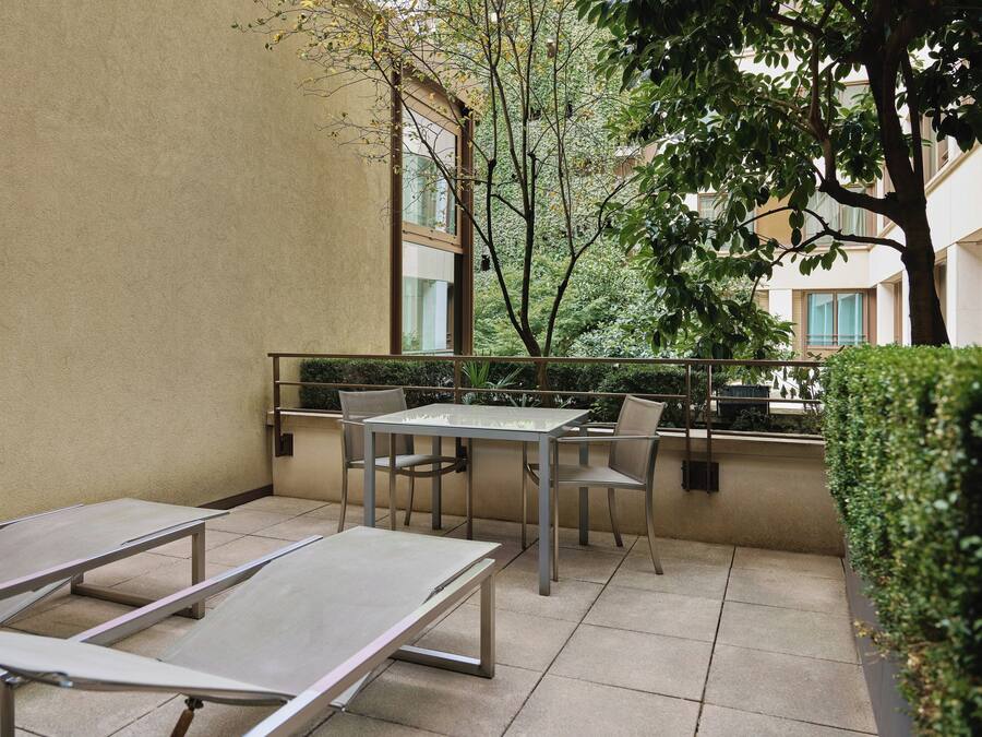 Uma varanda externa com mesa e cadeiras, um banco de metal e vista para um edifício e árvores.