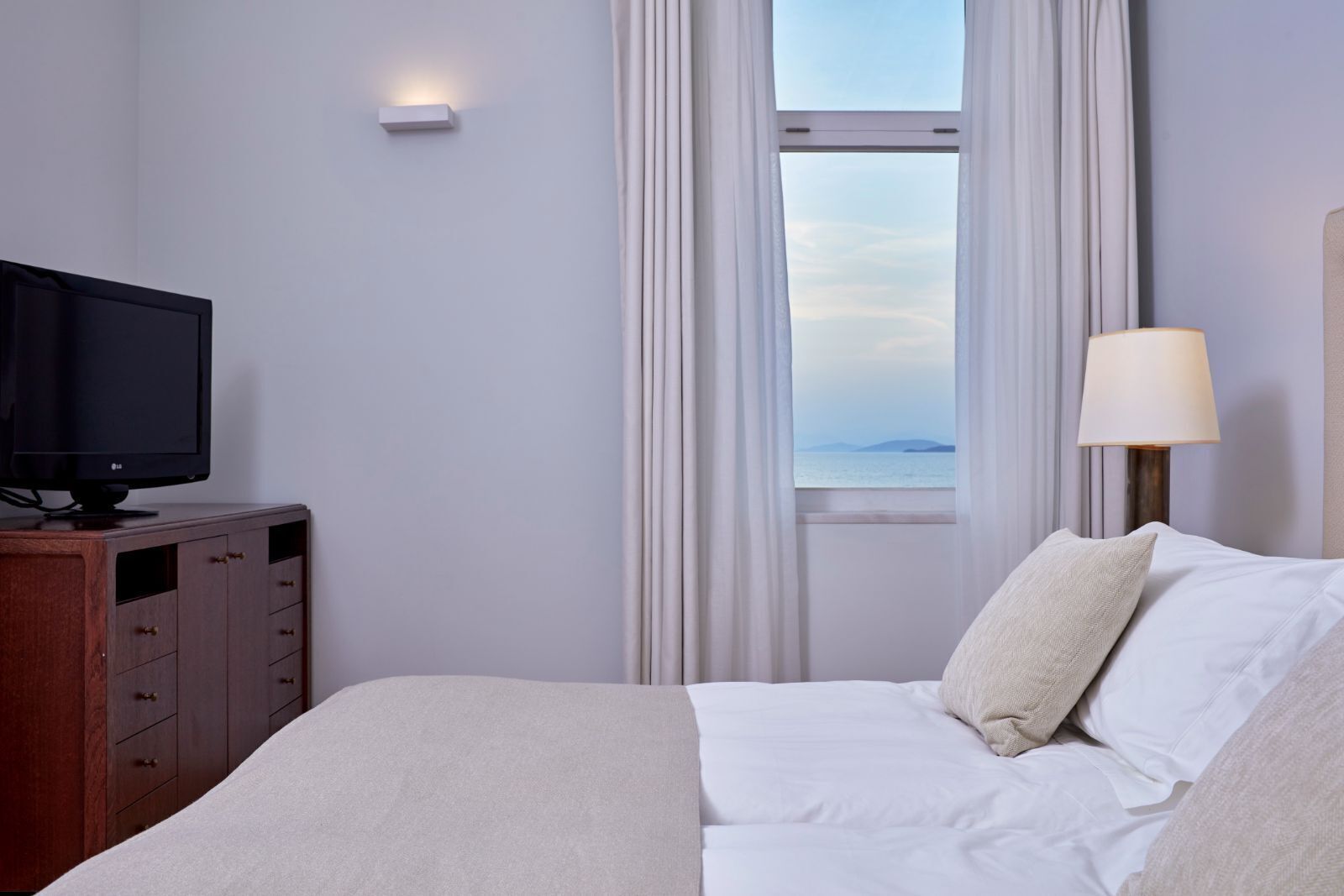 Quarto de hotel com cama, televisão em aparador de madeira, janela com cortinas e abajur.