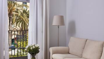 Sala de estar com sofá bege, luminária de chão, vaso com flores e vista para palmeiras através de uma janela com cortinas.