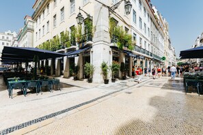 Rua com mesas ao ar livre, vasos de plantas e um edifício com várias lojas.
