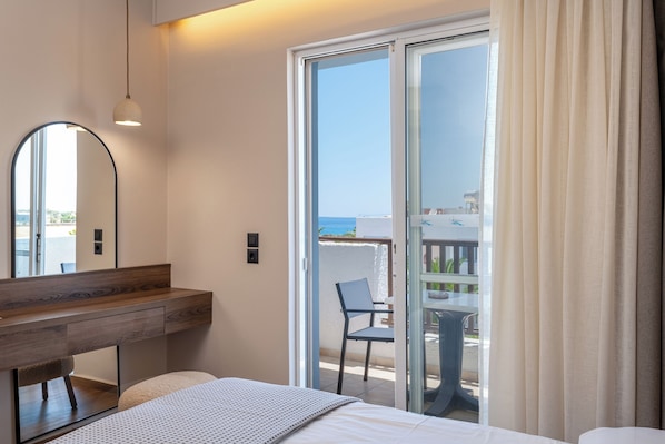 Quarto de hotel moderno com escrivaninha de madeira, espelho grande, cama e varanda com mesa e cadeiras.