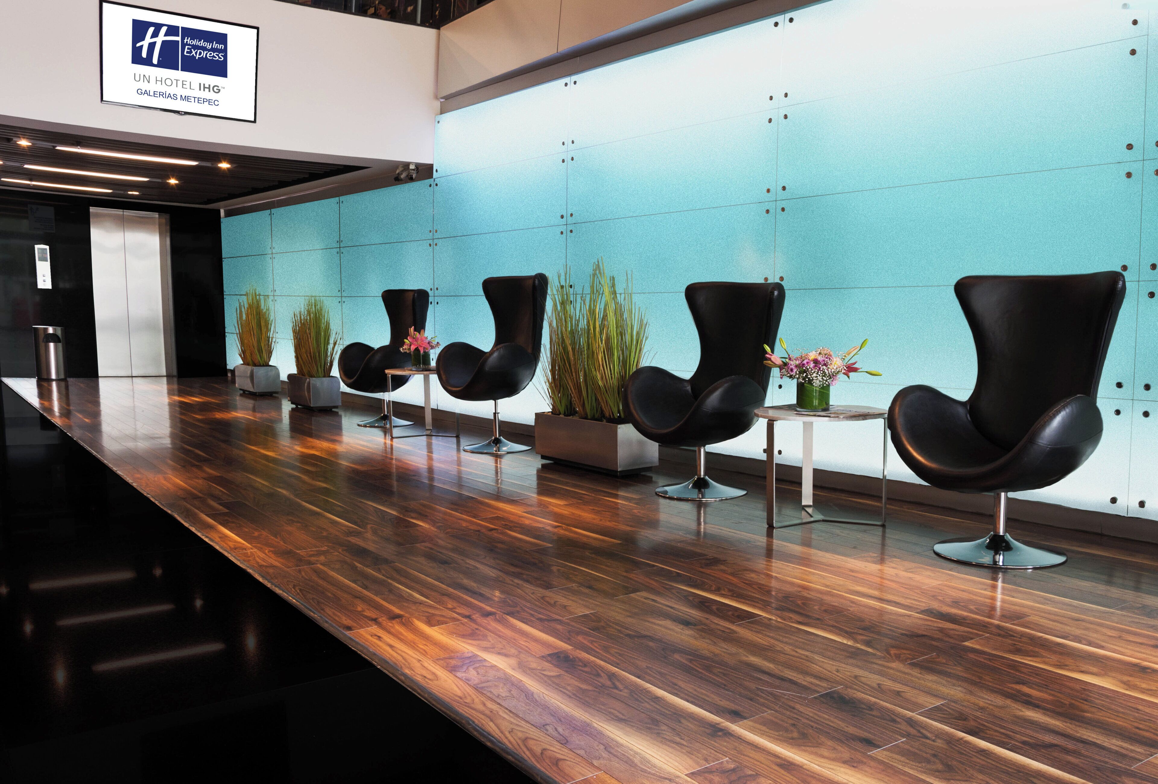 Uma área de recepção de hotel com design moderno, destacando um balcão de atendimento espaçoso, cadeiras estofadas em preto e plantas em vasos.