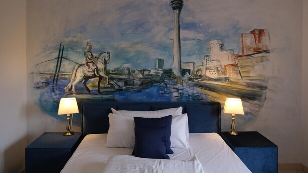 Quarto de hotel com uma cama, duas mesas de cabeceira e uma grande pintura de paisagem urbana na parede.