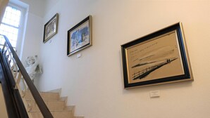 Uma escadaria com degraus bege e corrimão preto, que leva a um patamar com três obras de arte emolduradas na parede.