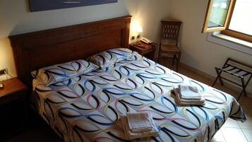 Uma cama bem arrumada com colcha estampada, cabeceira de madeira, mesa de cabeceira com abajur, uma cadeira e uma janela com vista.