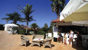 Uma área externa com assentos de vime, cadeiras e uma mesa, um bar com pessoas e palmeiras ao fundo.