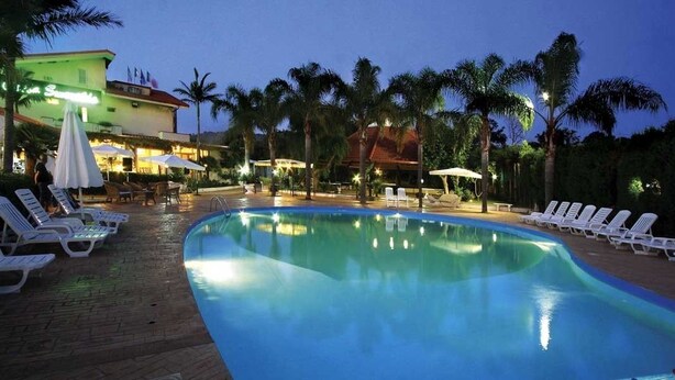 Piscina com espreguiçadeiras e guarda-sóis, cercada por palmeiras e um edifício com uma placa.