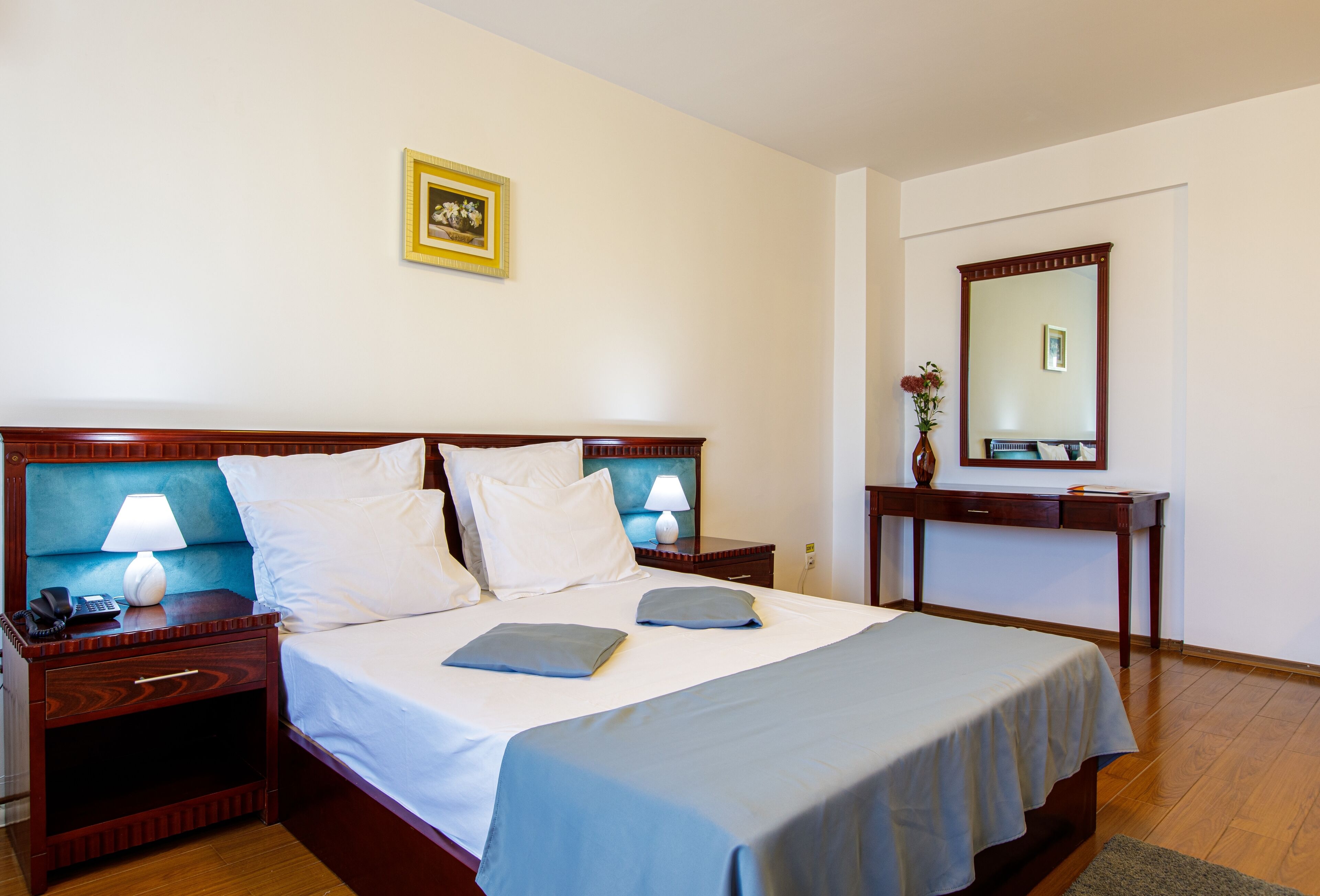 Quarto de hotel com cama, mesas de cabeceira, escrivaninha, espelho e quadro na parede.