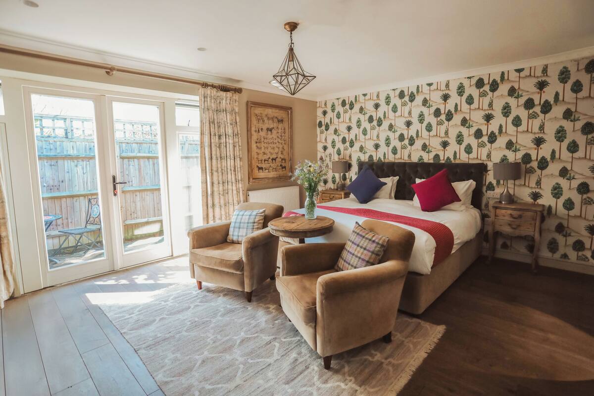 Quarto com uma cama grande, duas poltronas, uma mesa pequena com um vaso, uma mesa de cabeceira com abajur e vista para um deck de madeira através de uma porta de vidro.
