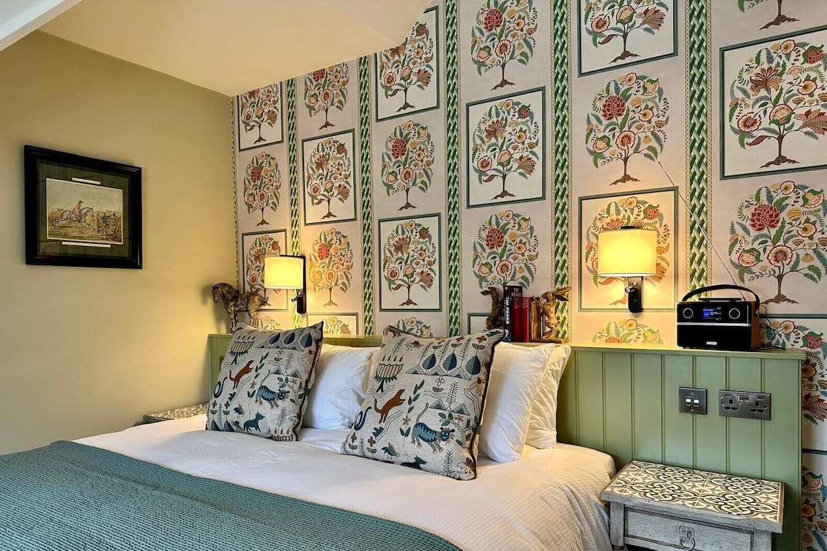 Quarto com cama, duas almofadas, mesa de cabeceira, abajur, rádio e um quadro na parede.