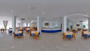 Ampla área de refeições de hotel, com mesas e cadeiras redondas, um balcão de buffet central e grandes janelas que proporcionam luz natural.