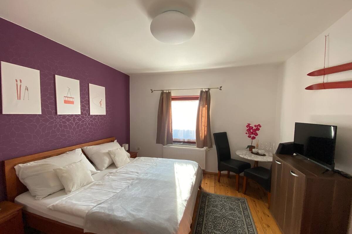 Quarto com cama, escrivaninha, televisão e janela com cortinas.