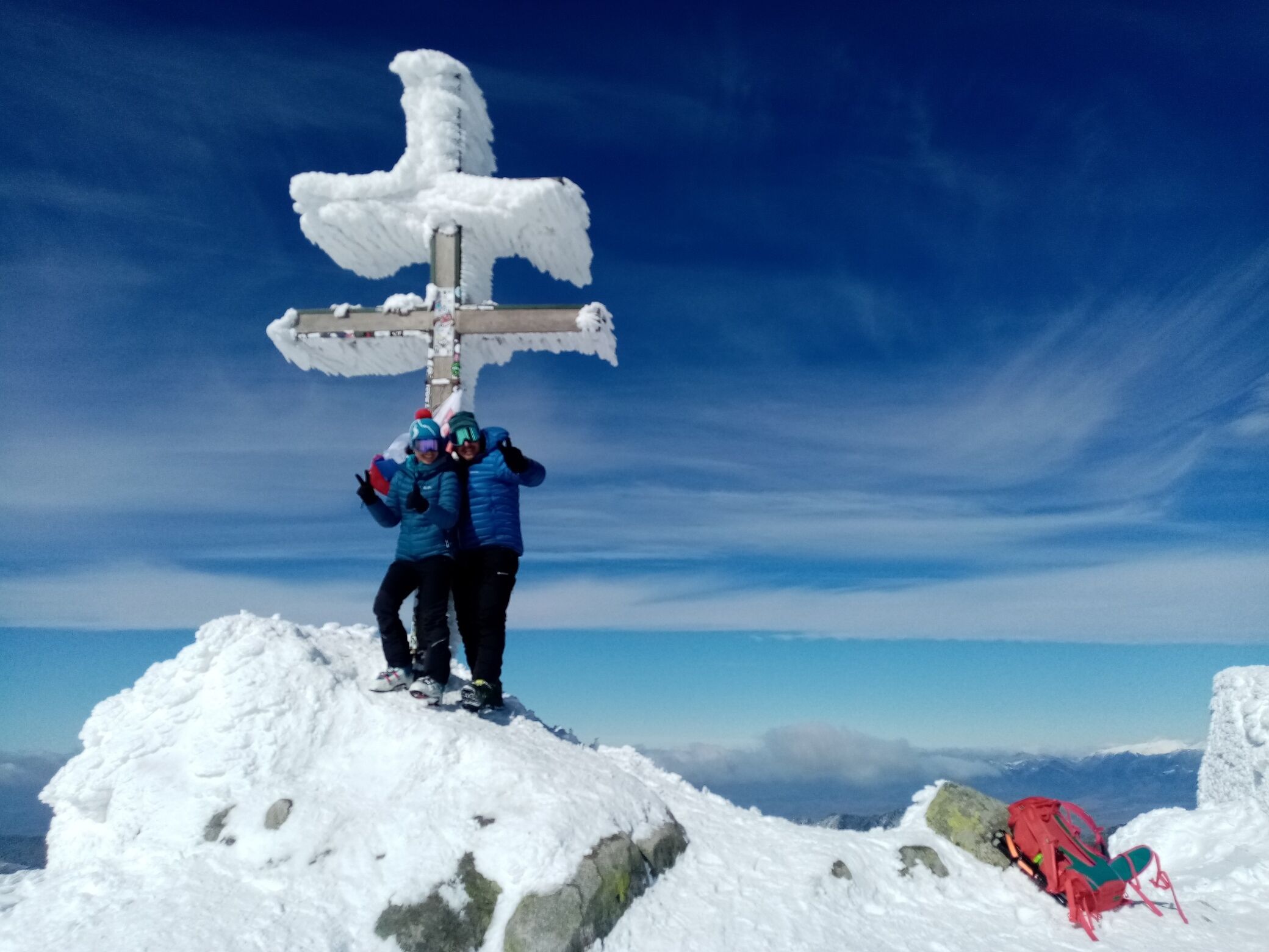 Pico nevado de montanha com uma escultura de neve em formato de cruz e uma mochila vermelha no chão.