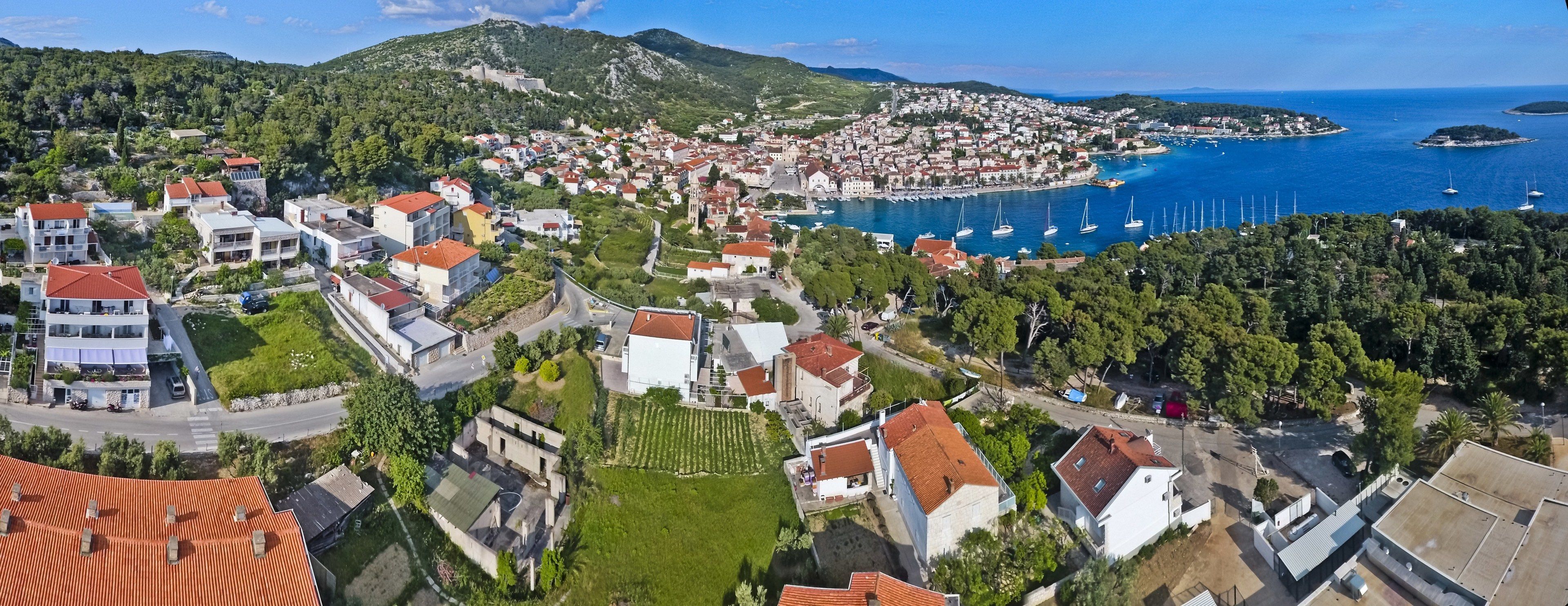 Vista aérea de uma vila costeira com mistura de edifícios residenciais e comerciais, um porto com barcos e uma colina imponente com uma fortaleza.