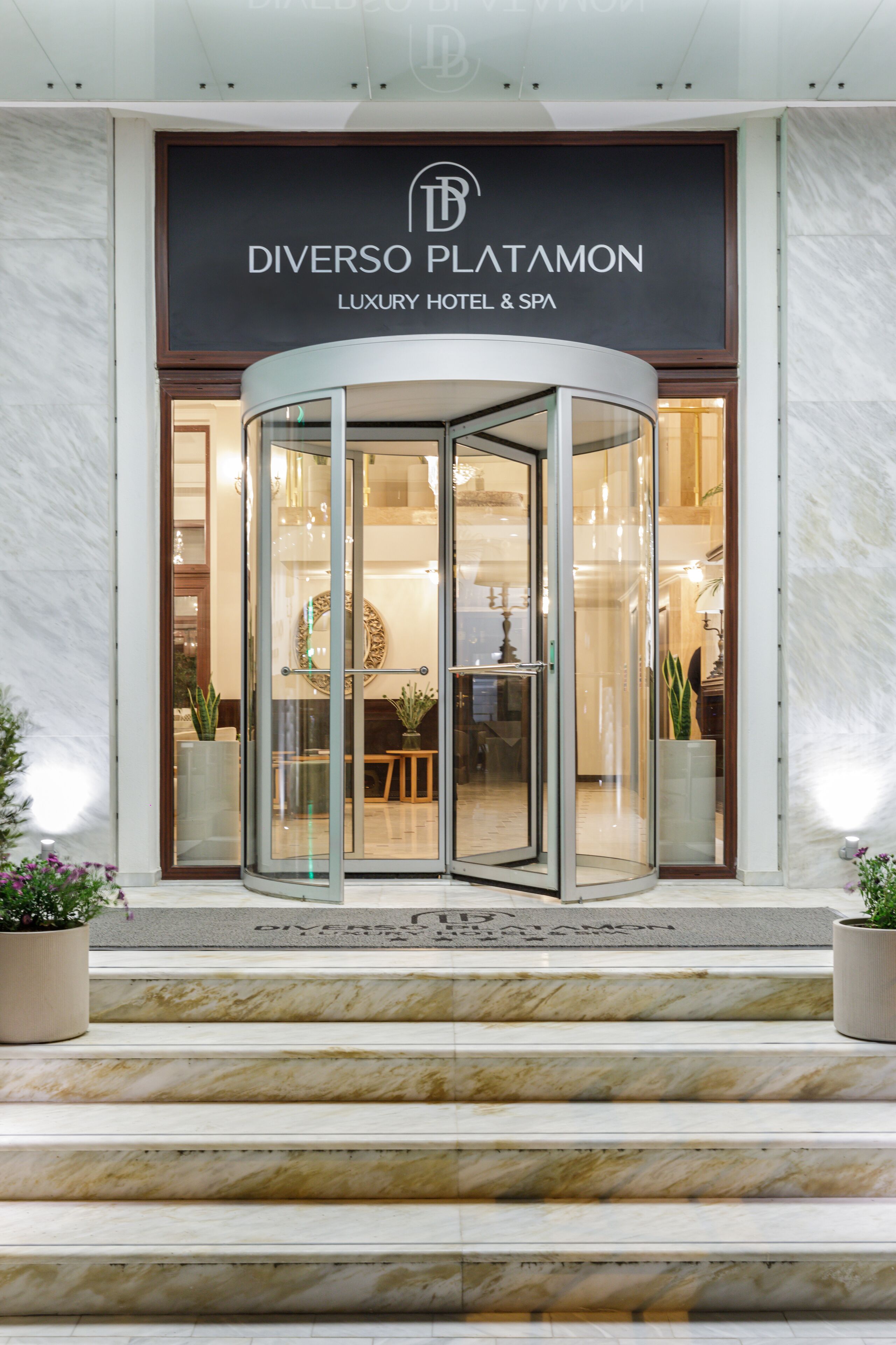 Entrada de um hotel de luxo com porta giratória, degraus de mármore e uma placa com a inscrição "DIVERSO PLATAMON LUXURY HOTEL & SPA".
